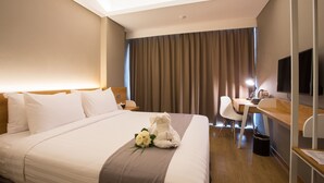 Quarto de hotel com cama bem arrumada, escrivaninha com cadeira, televisão e janela com cortinas.
