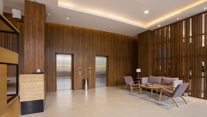 Um lobby moderno com painéis de madeira, um balcão de recepção, área de estar com sofá e mesa de centro, e dois elevadores.