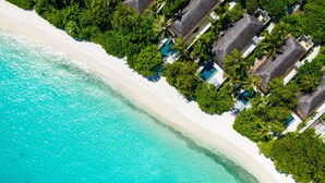 Vista aérea de um resort à beira-mar, com areia branca, água turquesa e edifícios com telhados de palha.