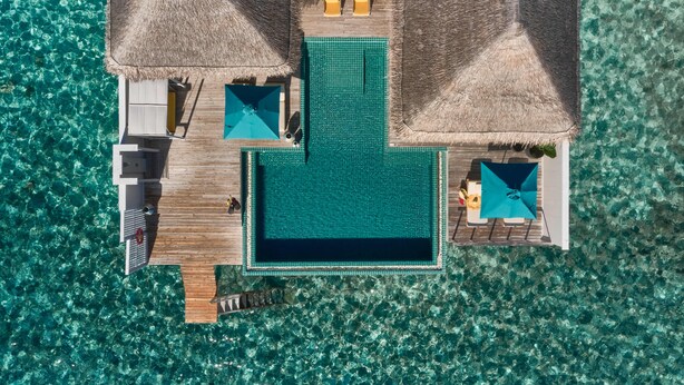 Vista aérea de um resort com piscina central, duas estruturas com telhados de palha e espreguiçadeiras.