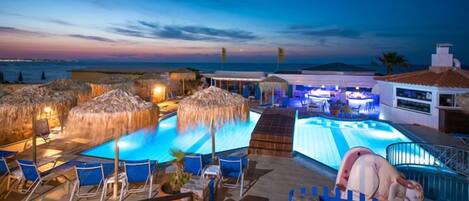 Área da piscina com espreguiçadeiras e bar, com vista para um corpo de água.