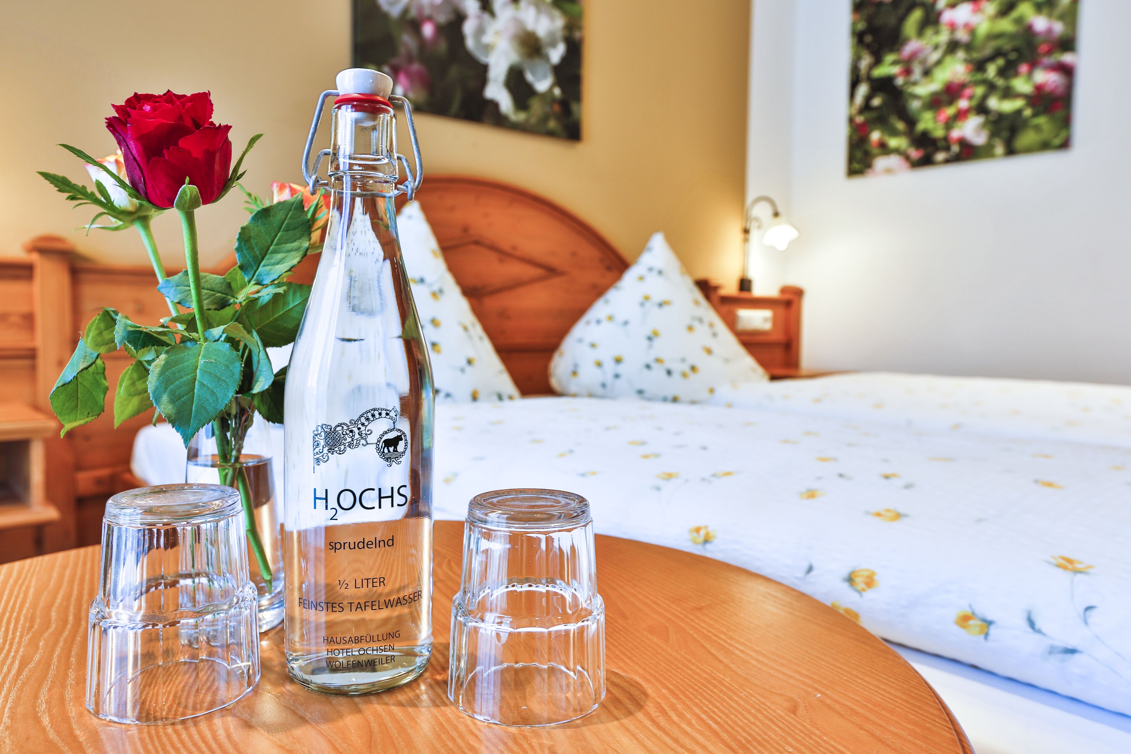 Uma garrafa de Hockspund sprudel, dois copos e uma rosa vermelha sobre uma mesa de madeira num quarto de hotel com cama e decoração floral na parede.