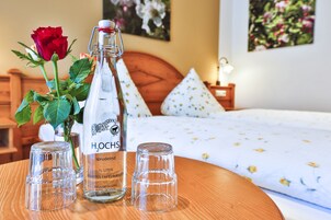 Uma garrafa de Hockspund sprudel, dois copos e uma rosa vermelha sobre uma mesa de madeira num quarto de hotel com cama e decoração floral na parede.