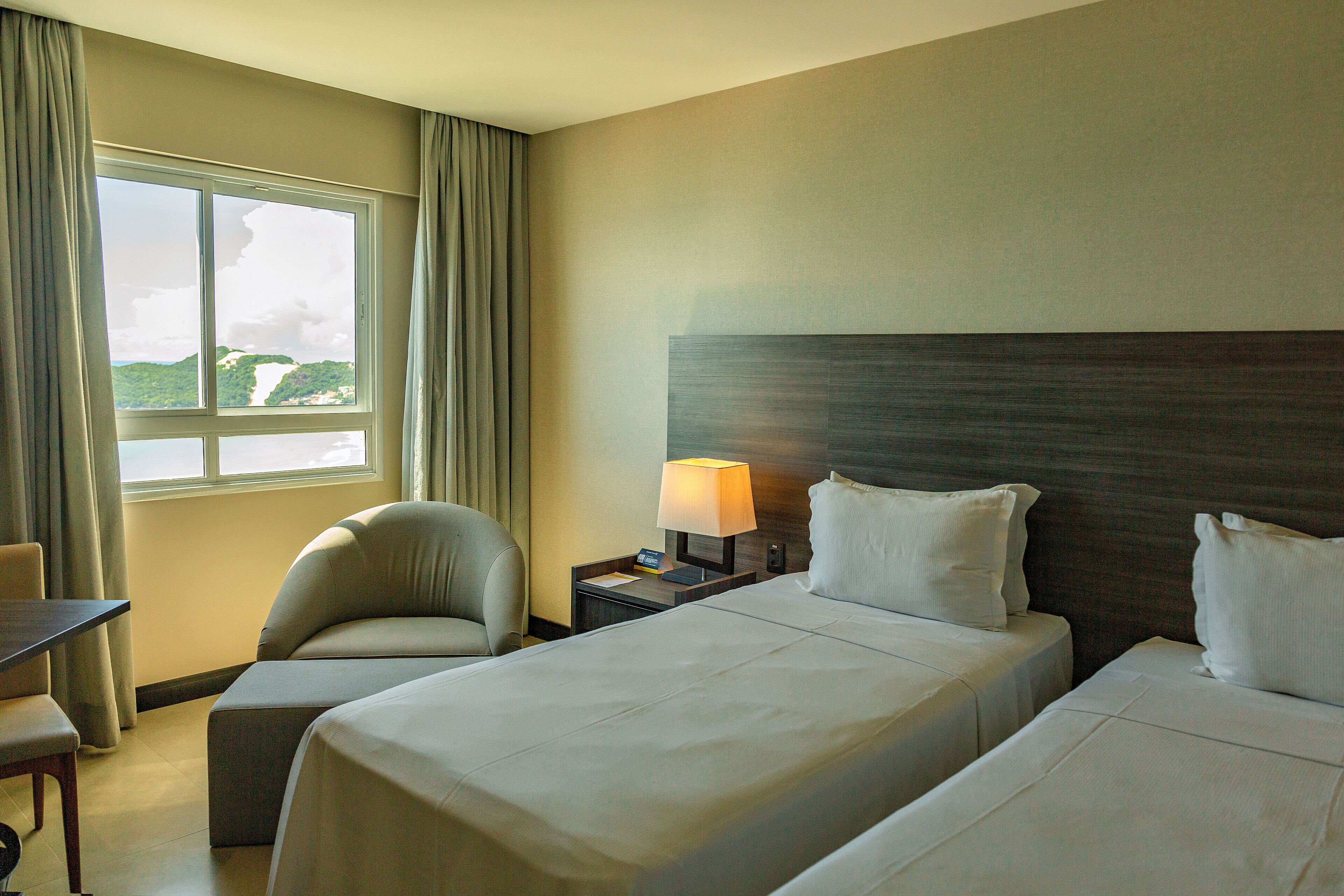 Quarto de hotel com duas camas, uma escrivaninha, uma cadeira e janela com vista para a paisagem.