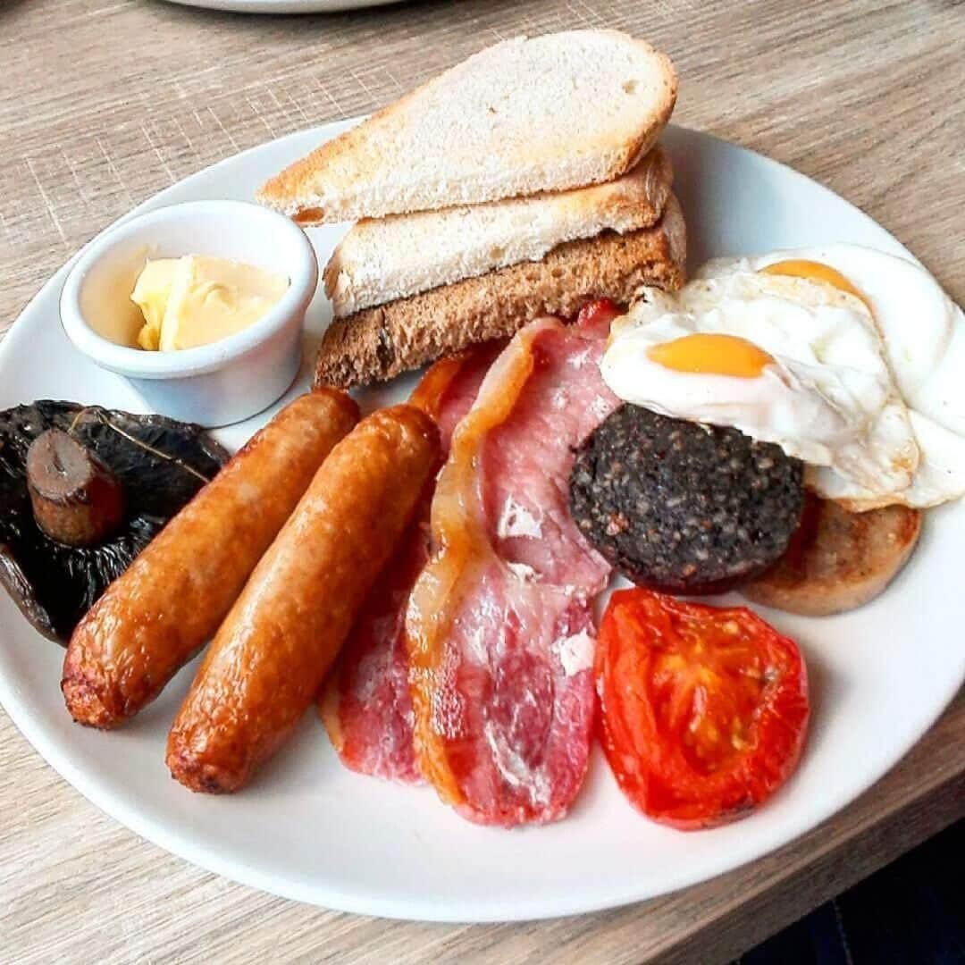 Um prato de comida de café da manhã com salsichas, bacon, ovos, torradas e uma pequena tigela de manteiga.