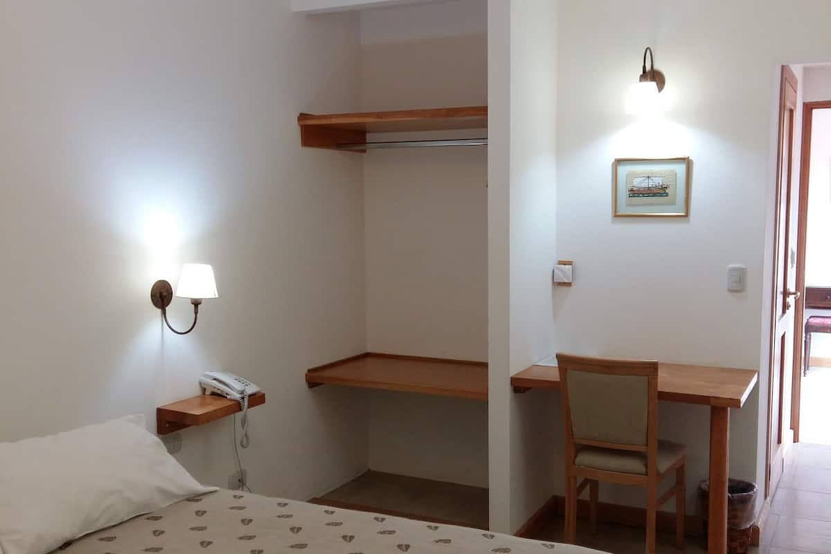 Quarto com cama, mesa de cabeceira com abajur, escrivaninha com cadeira, prateleira e quadro na parede.