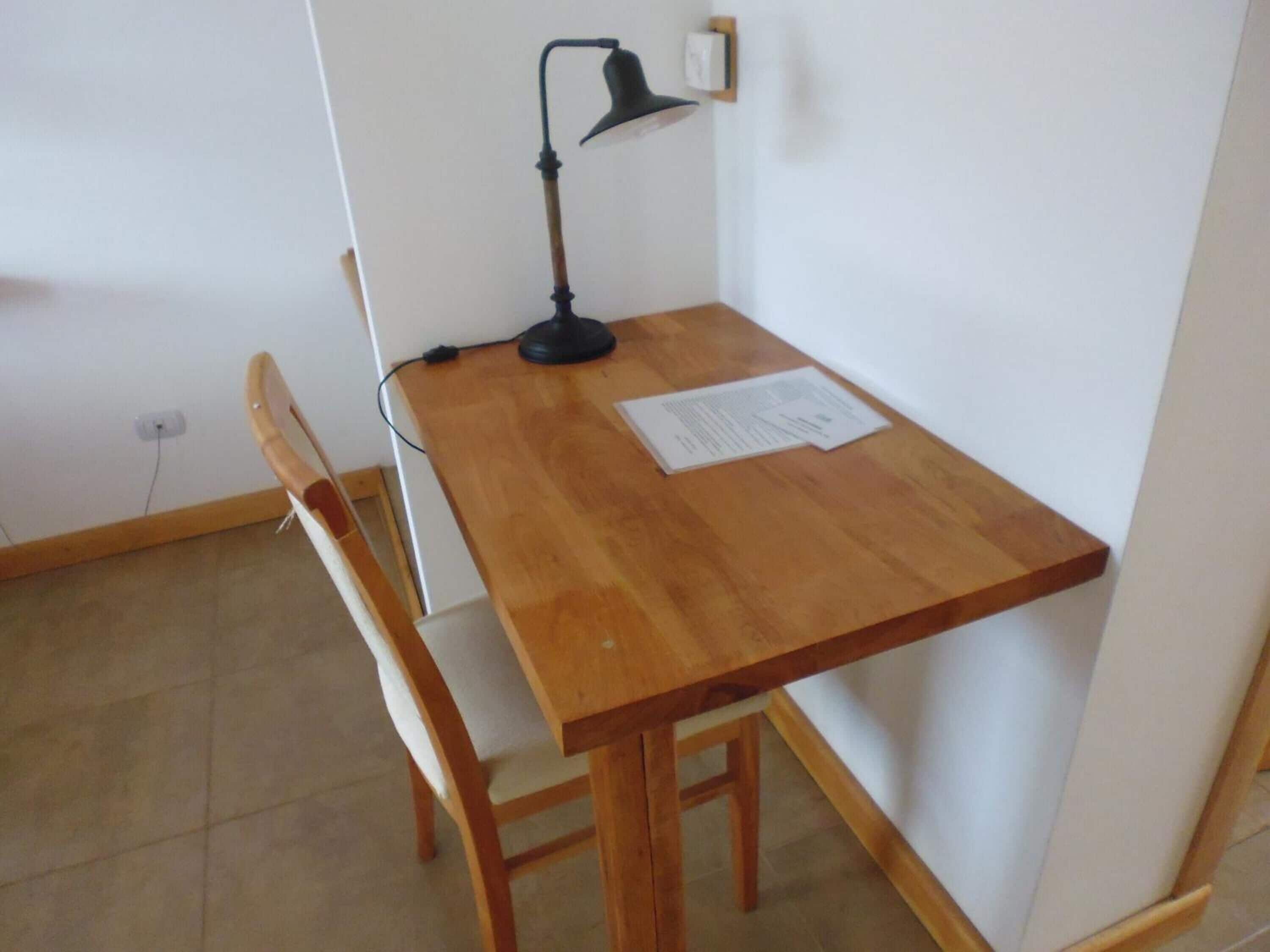 Uma mesa de madeira com uma luminária de mesa preta, papéis e uma cadeira em um quarto com paredes brancas e piso ladrilhado.