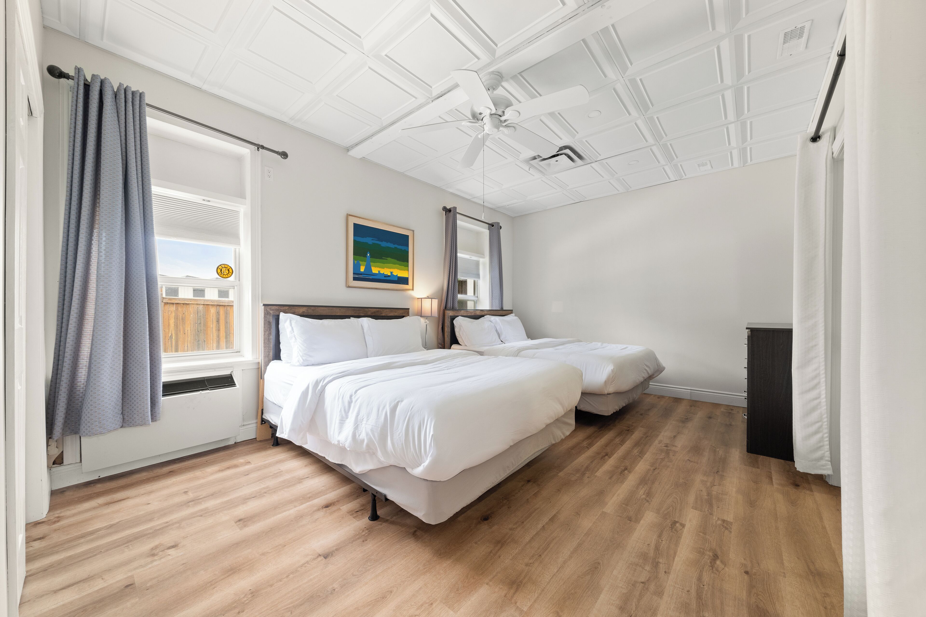 Um quarto com duas camas, um ventilador de teto, uma janela com cortinas e um quadro na parede.