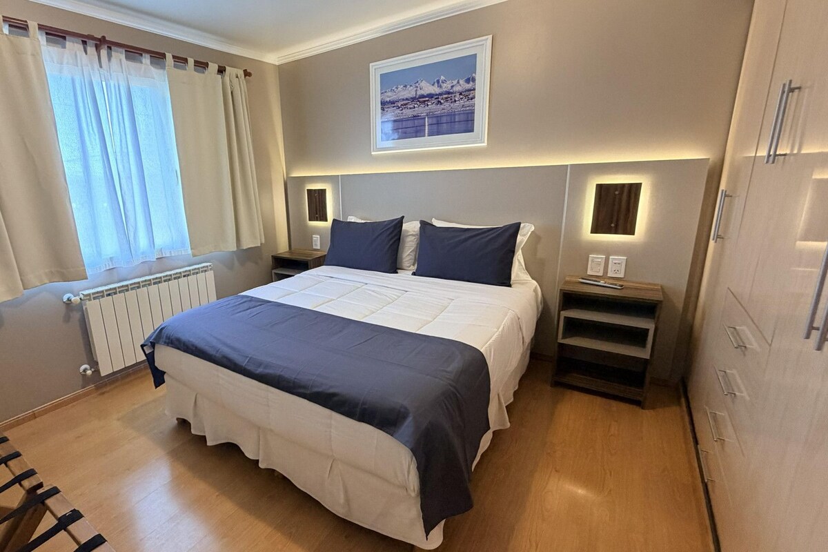Quarto com cama, janela com cortinas, mesa de cabeceira, radiador e quadro na parede.