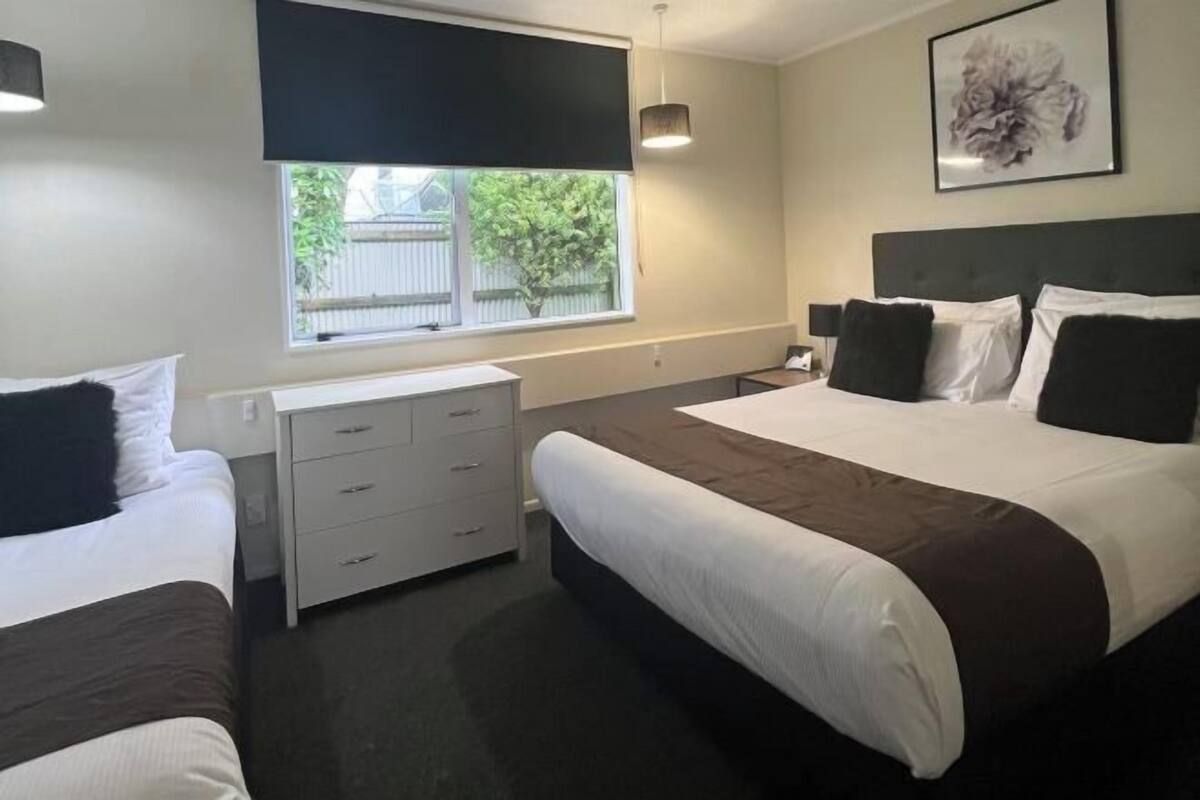 Quarto de hotel com duas camas, uma secretária, uma cadeira, janela com vista para vegetação e um quadro na parede.
