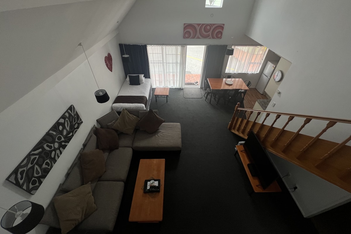 Uma sala moderna com um sofá, mesa de centro e uma área de jantar com mesa e cadeiras. Existe uma escada que leva a um andar superior.