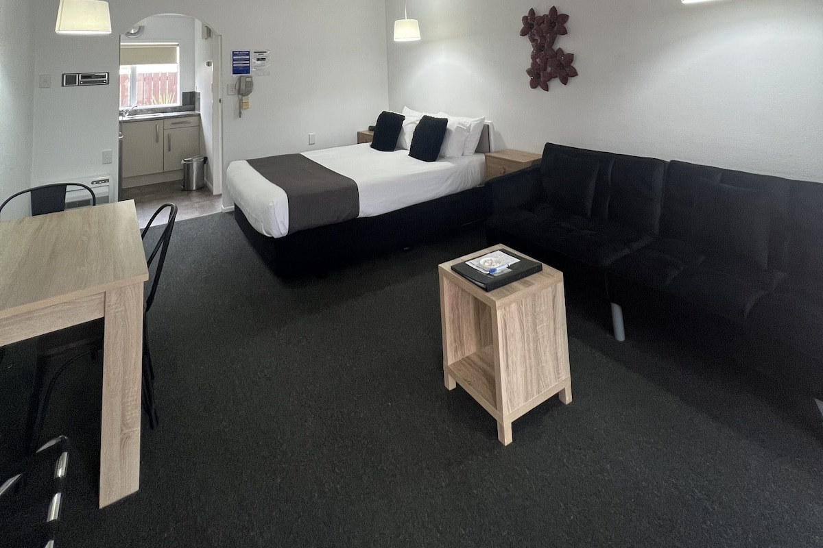 Quarto de hotel compacto com uma cama, um sofá preto, uma pequena mesa de madeira e uma cozinha compacta ao fundo.
