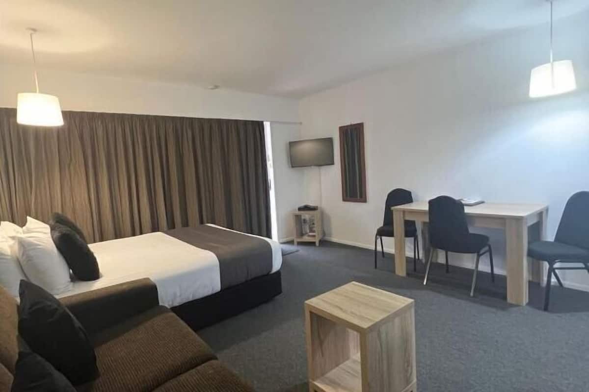 Quarto de hotel com uma cama, uma escrivaninha, uma cadeira, uma televisão e uma mesinha.