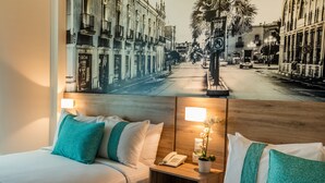 Quarto de hotel com duas camas, uma mesa de cabeceira com telefone e um vaso de planta, e uma janela grande com vista para a rua da cidade.
