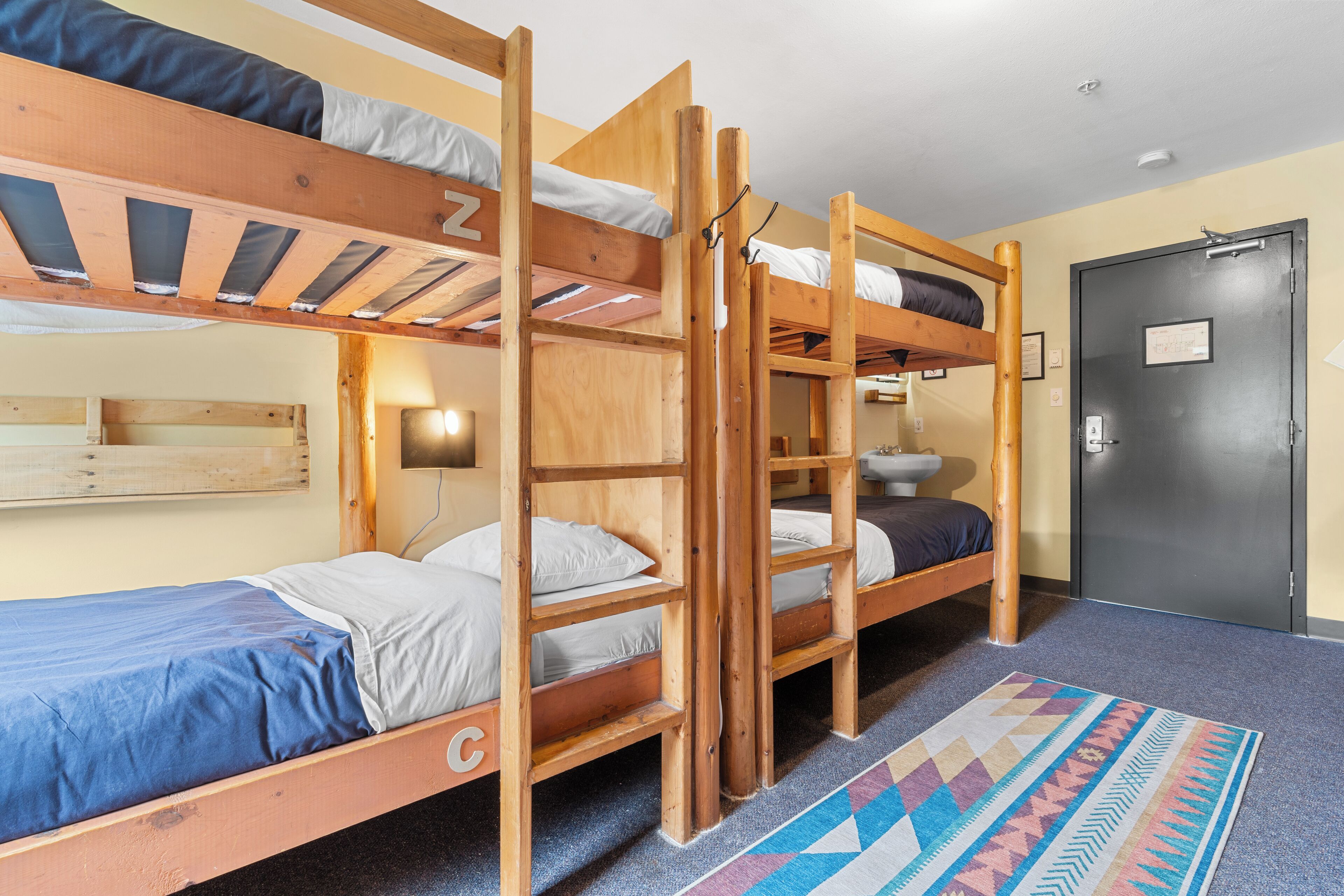 Quarto com beliche de madeira, uma cama simples, banheiro com pia e porta com aviso.