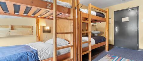 Quarto com beliche de madeira, uma cama simples, banheiro com pia e porta com aviso.
