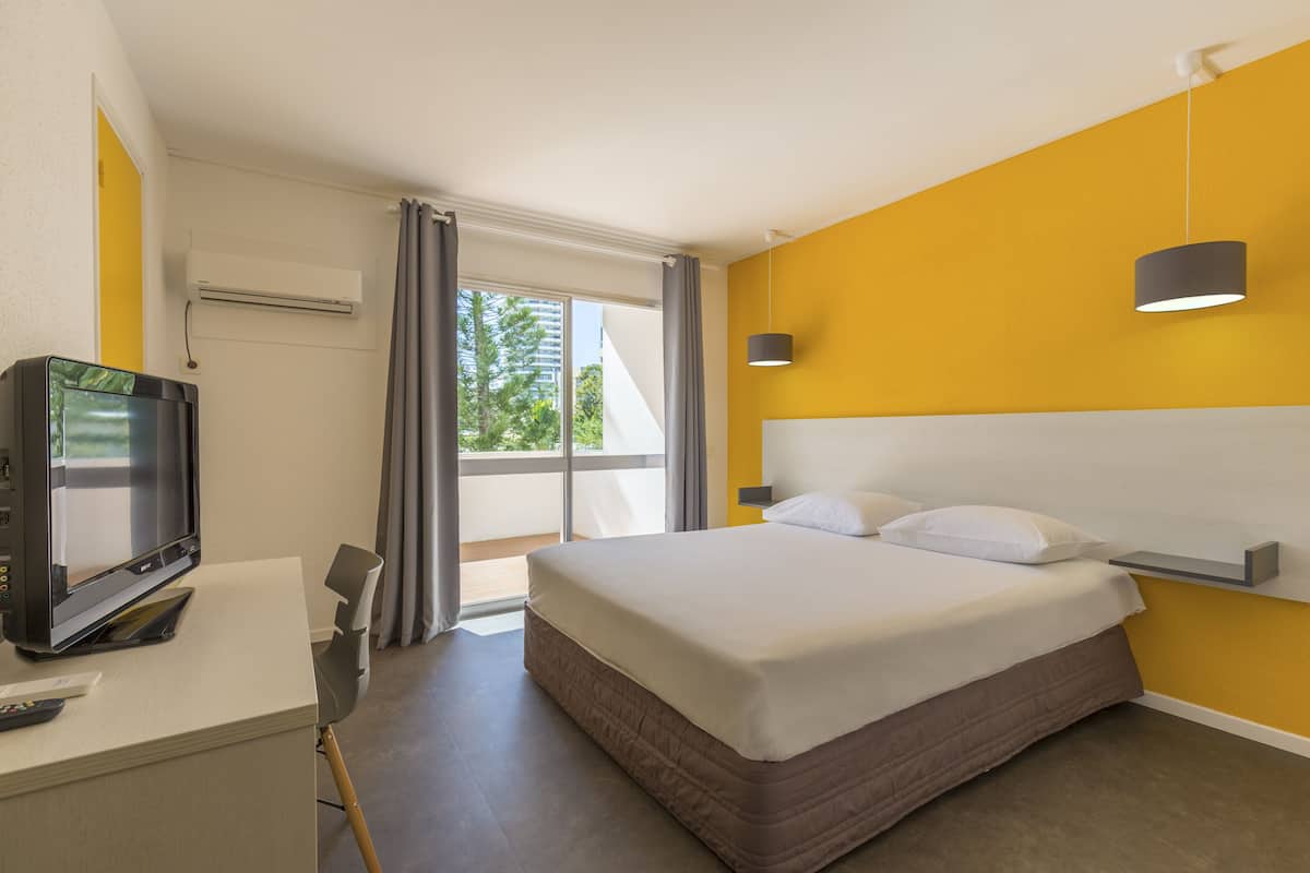 Quarto de hotel com cama, escrivaninha com televisão e janela com cortinas.