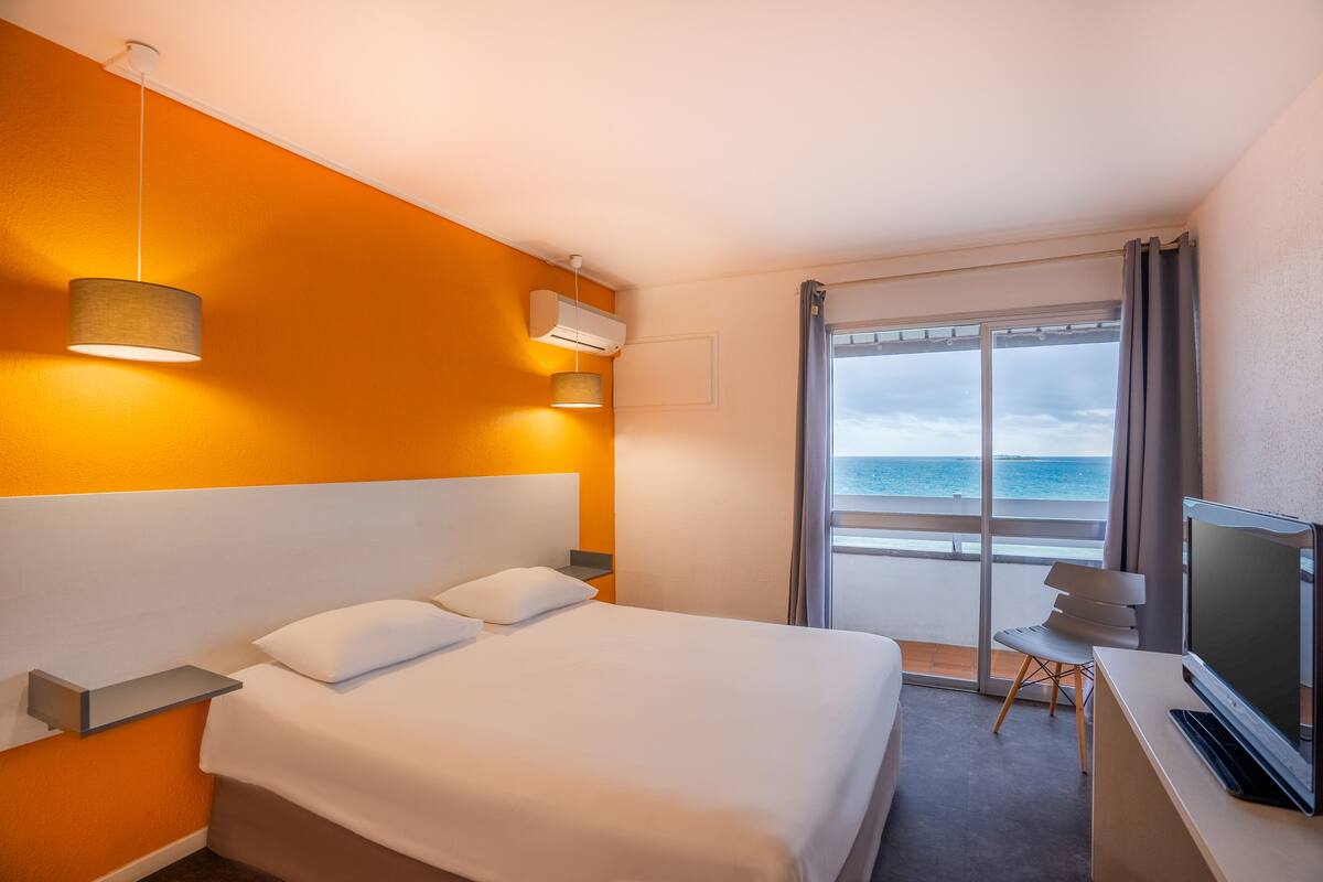 Quarto de hotel com cama, escrivaninha com televisão e vista para o mar através de uma grande janela.