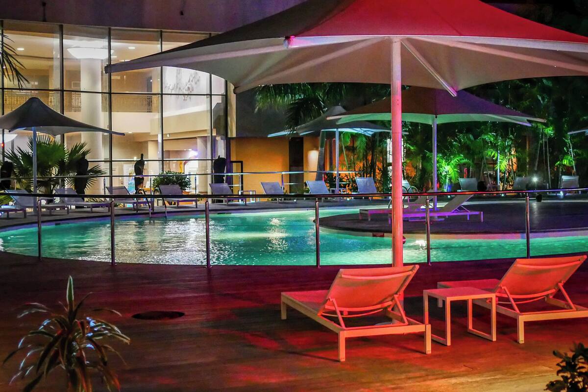 Uma área de piscina com espreguiçadeiras e guarda-sóis, rodeada por um edifício com vários andares e janelas.