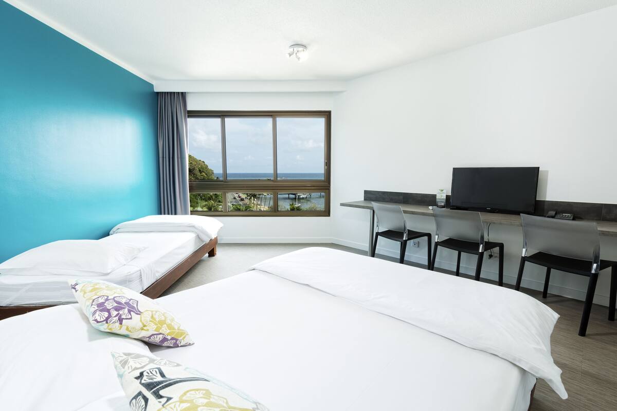 Quarto de hotel com duas camas, uma escrivaninha com televisão e uma janela grande com vista para o mar.