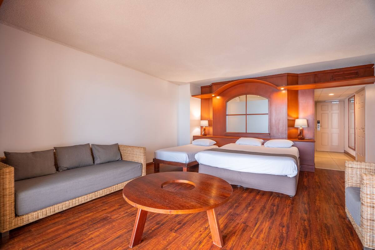 Quarto de hotel com sofá, duas camas individuais, mesa redonda de madeira e espelho sobre as camas.
