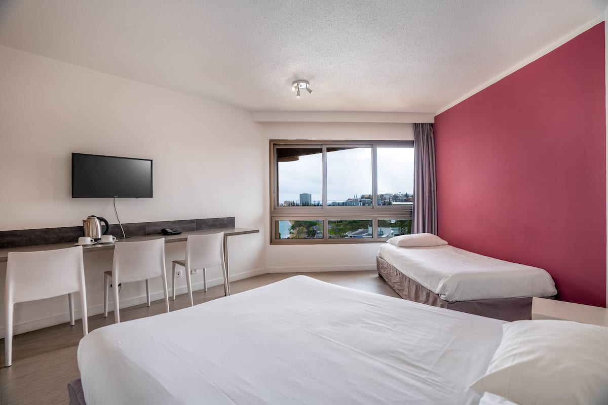 Quarto de hotel com cama, uma escrivaninha com cadeiras, televisão e uma janela grande com vista para a cidade.