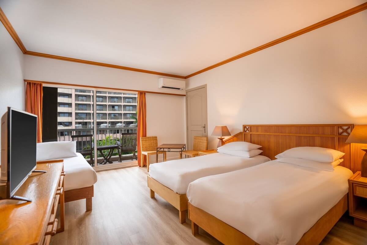 Quarto de hotel com duas camas, televisão, varanda com vista para um edifício e uma mesa com cadeiras.