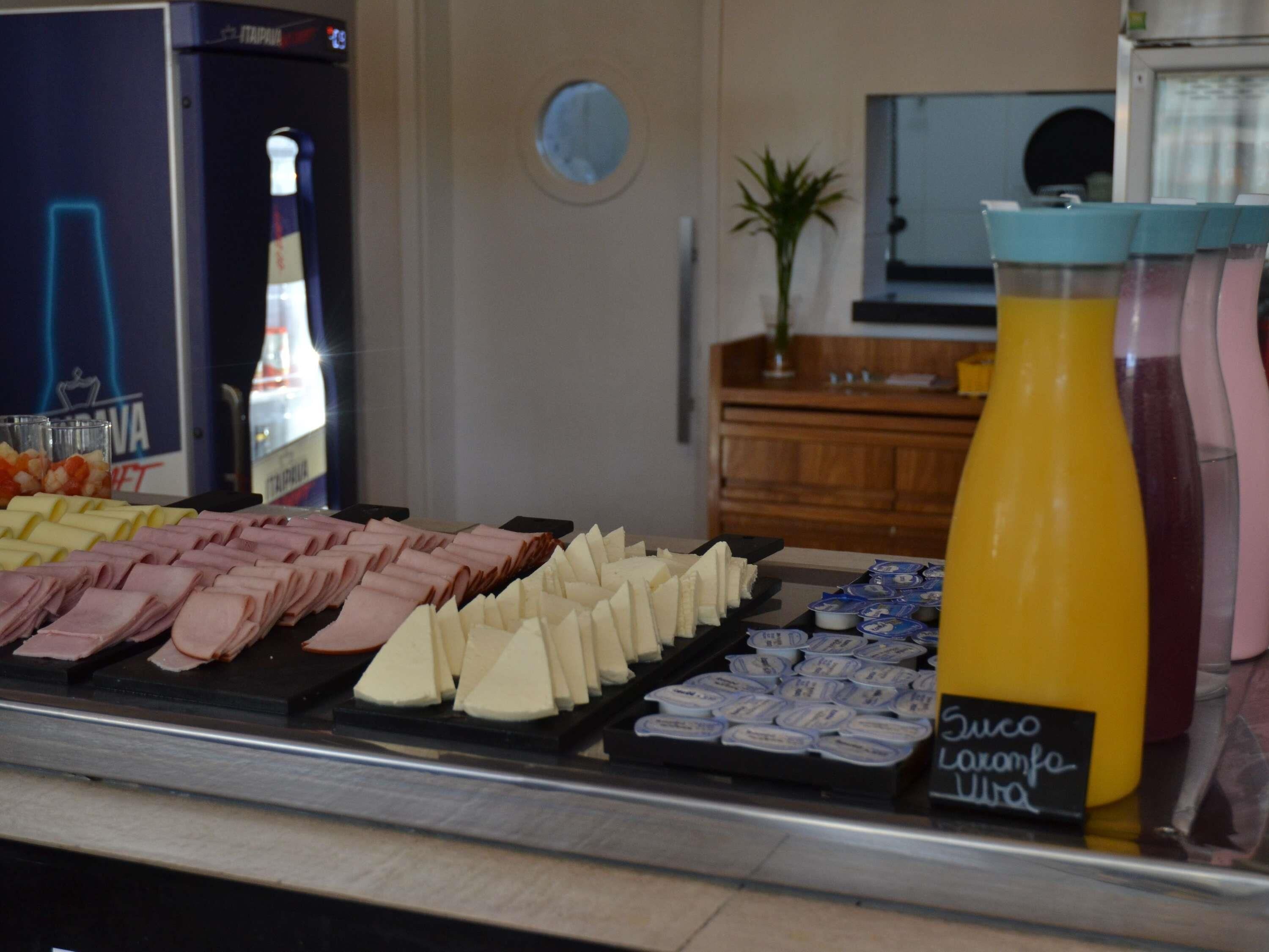 Buffet com diversos queijos e carnes, um dispensador de suco e um aviso sobre uma oferta especial.