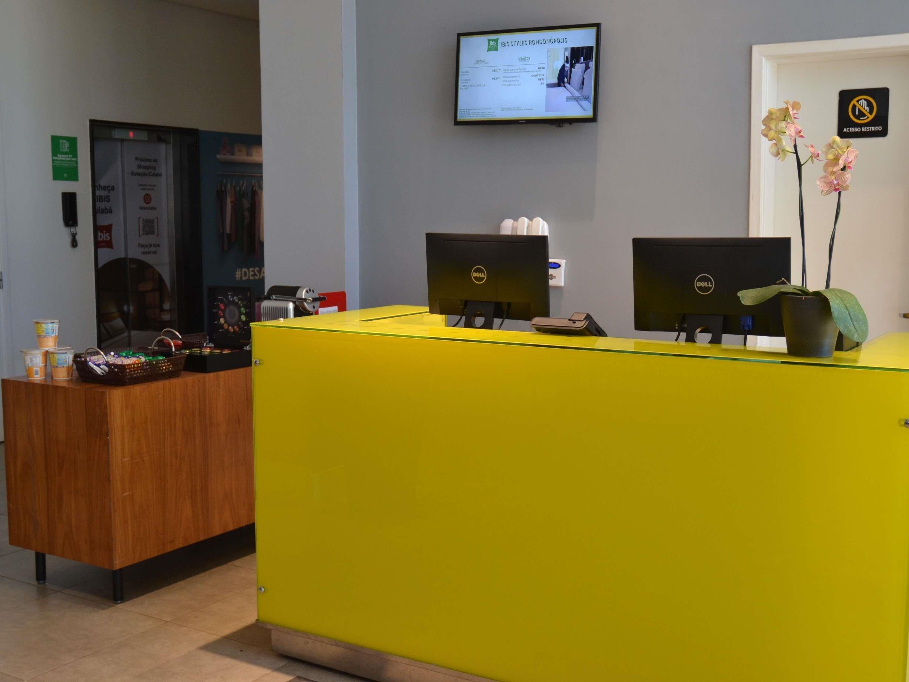 Uma área de recepção com uma mesa de madeira e um balcão amarelo, dois computadores Dell, uma televisão na parede e uma placa indicando acesso restrito.