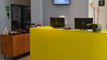 Uma área de recepção com uma mesa de madeira e um balcão amarelo, dois computadores Dell, uma televisão na parede e uma placa indicando acesso restrito.