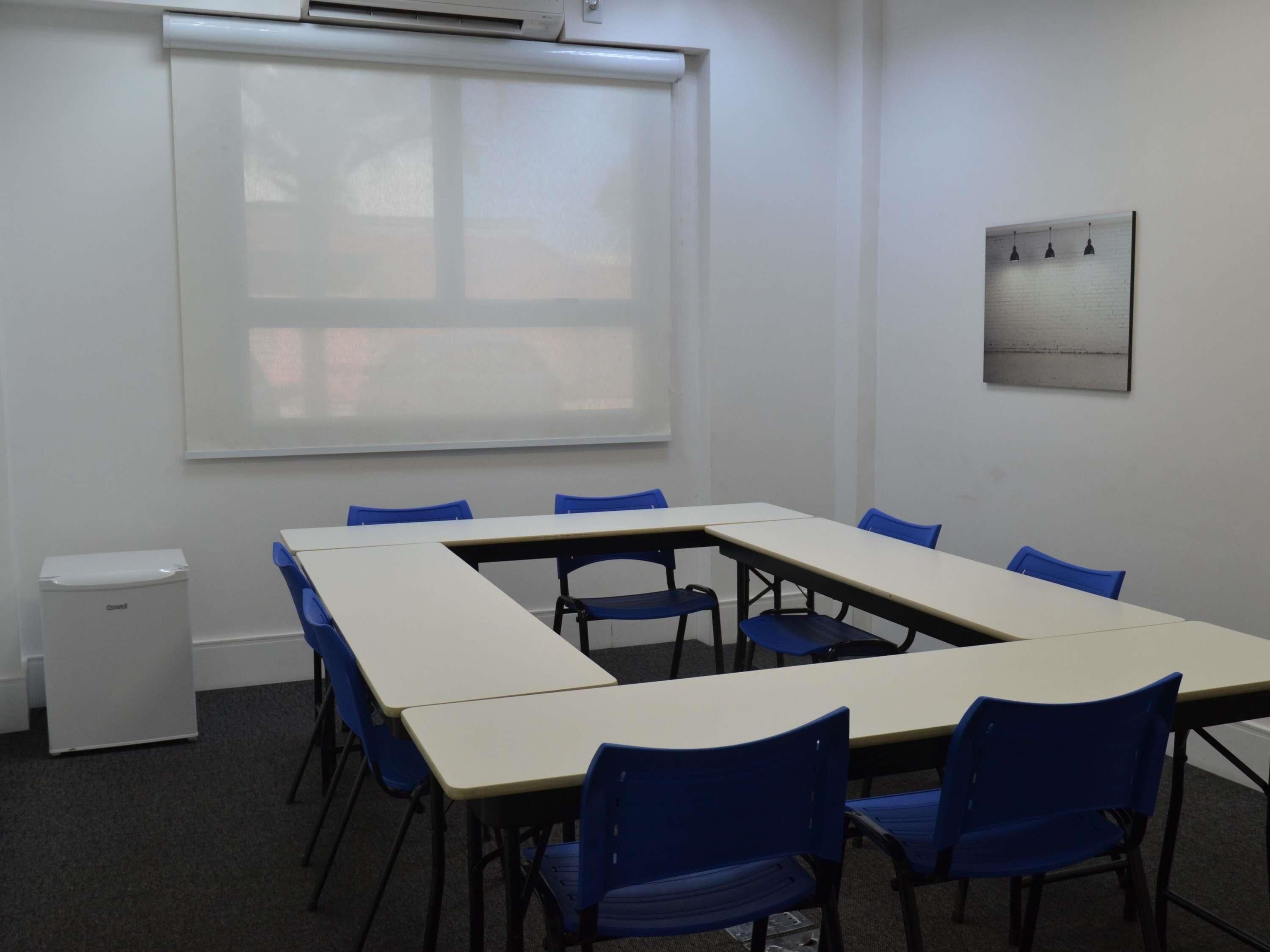 Uma sala de conferências com uma mesa grande, cadeiras azuis, uma tela de projetor e uma pequena lixeira.