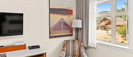 Quarto de hotel com cama, escrivaninha com TV, cadeira, pintura e janela com vista para um prédio e árvores.