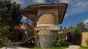 Um edifício com telhado de palha e uma escadaria que leva a uma varanda, rodeado por vegetação tropical.