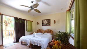 Quarto com cama, ventoinha no teto, janela com cortinas, pintura na parede e uma tigela de frutas sobre uma mesa.
