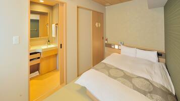 Quarto de hotel com cama, guarda-roupa de madeira, banheiro com espelho e prateleira pequena.