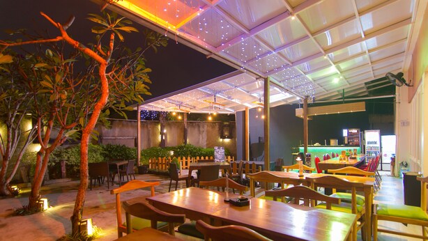 Uma área de refeições ao ar livre com mesas e cadeiras de madeira, um bar com balcão e banquetas, e uma seção coberta com luzes de Natal.