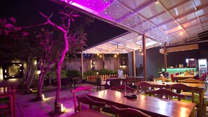 Uma área de refeições ao ar livre com mesas e cadeiras de madeira, uma pérgola com luzes e um bar ao fundo.