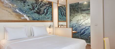 Quarto de hotel moderno com uma cama grande, cabeceira de madeira, espelho fixado na parede e banheiro com box de vidro.