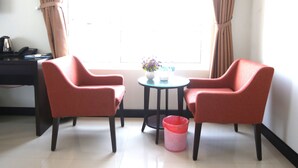Quarto com duas cadeiras vermelhas, uma mesinha com um vaso de flores, uma lixeira vermelha e uma escrivaninha com chaleira e telefone.