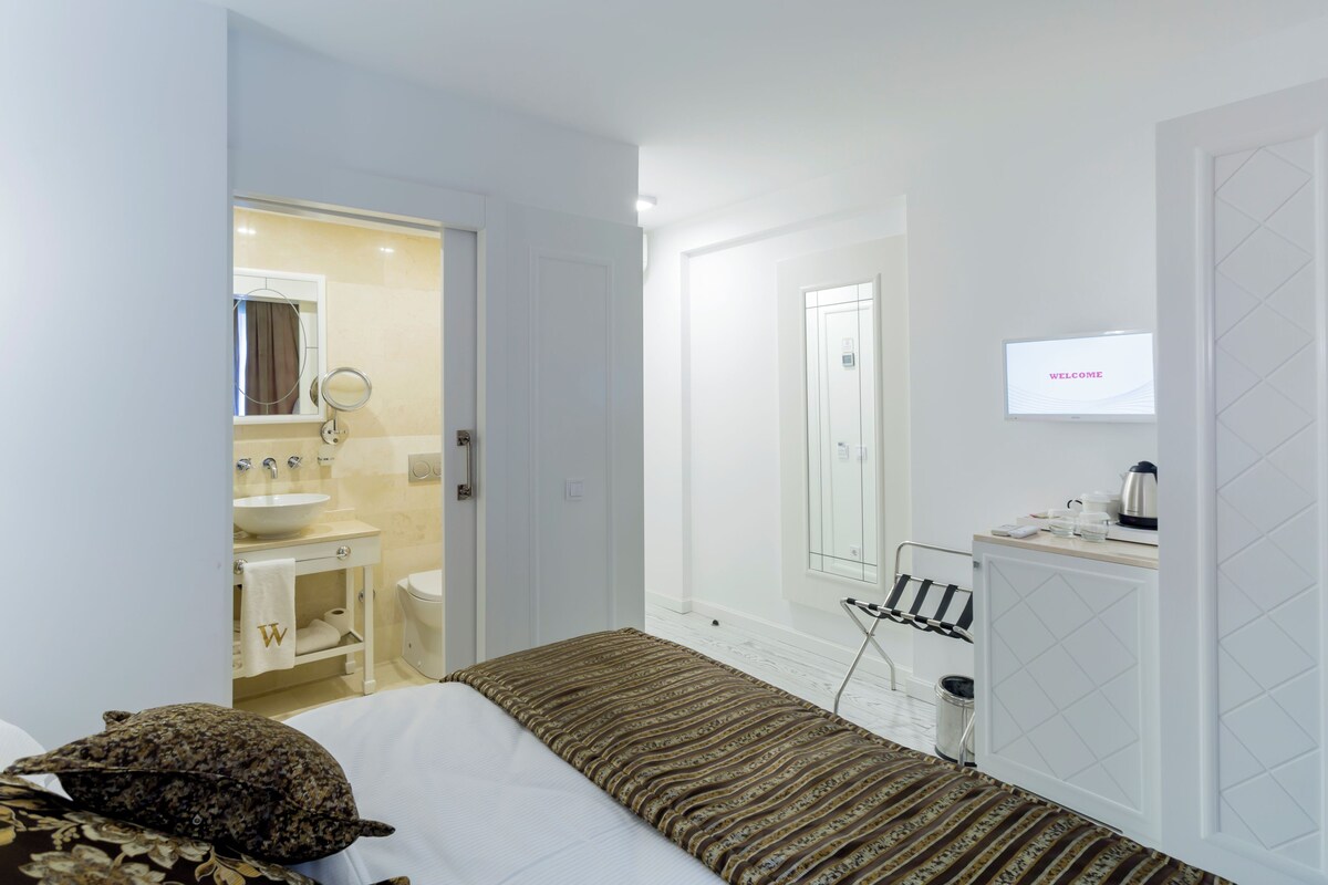 Quarto de hotel com cama, mesa de cabeceira, televisão, banheiro com pia e vaso sanitário, e chuveiro.