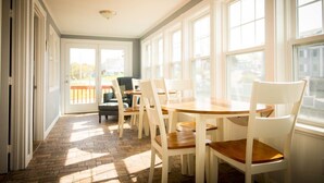 Quarto iluminado pelo sol com uma mesa de jantar e cadeiras, janelas amplas e vista para o exterior.