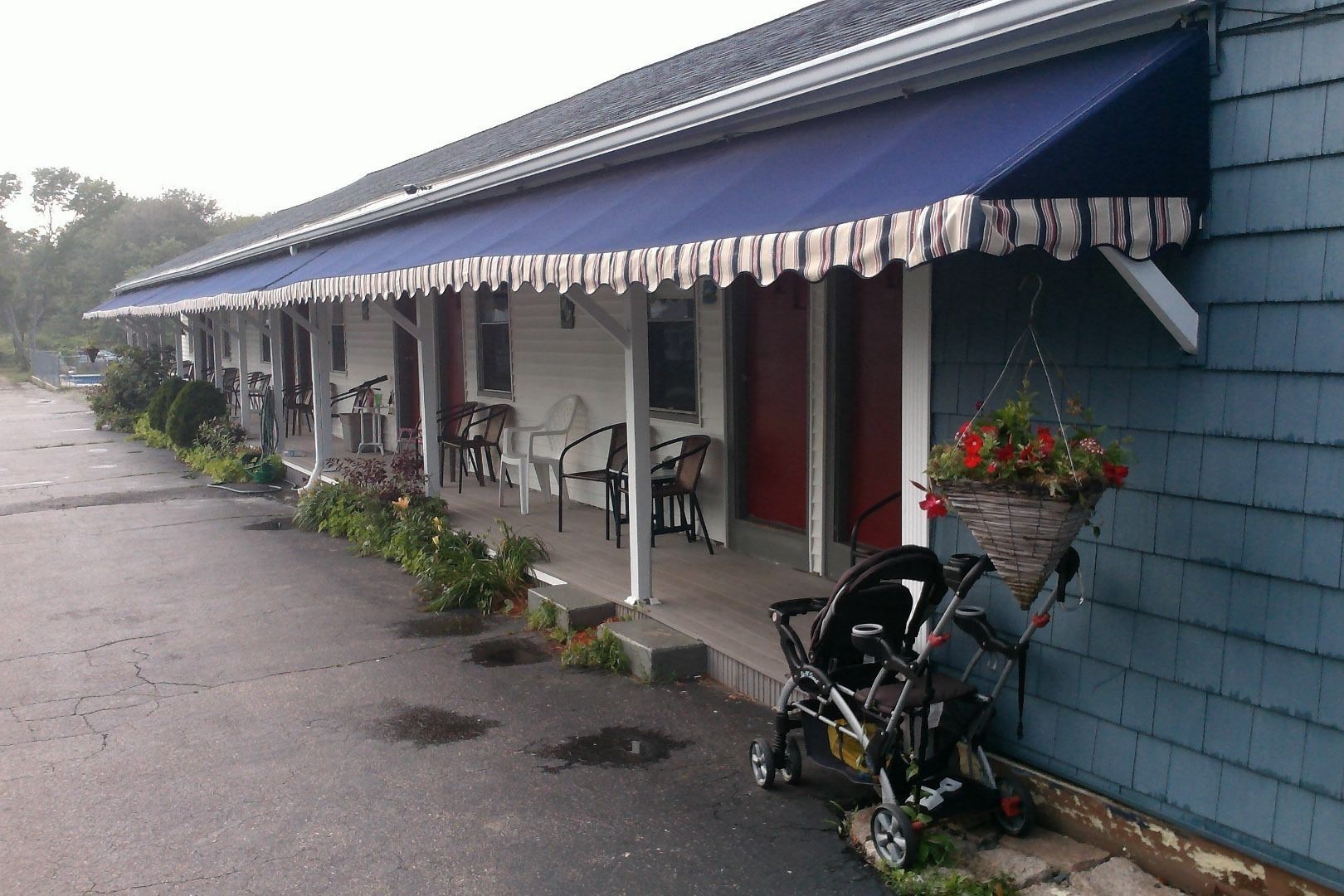 Uma fileira de quartos de motel, cada um com toldo individual, área de estar externa e um carrinho de bebê estacionado na frente.