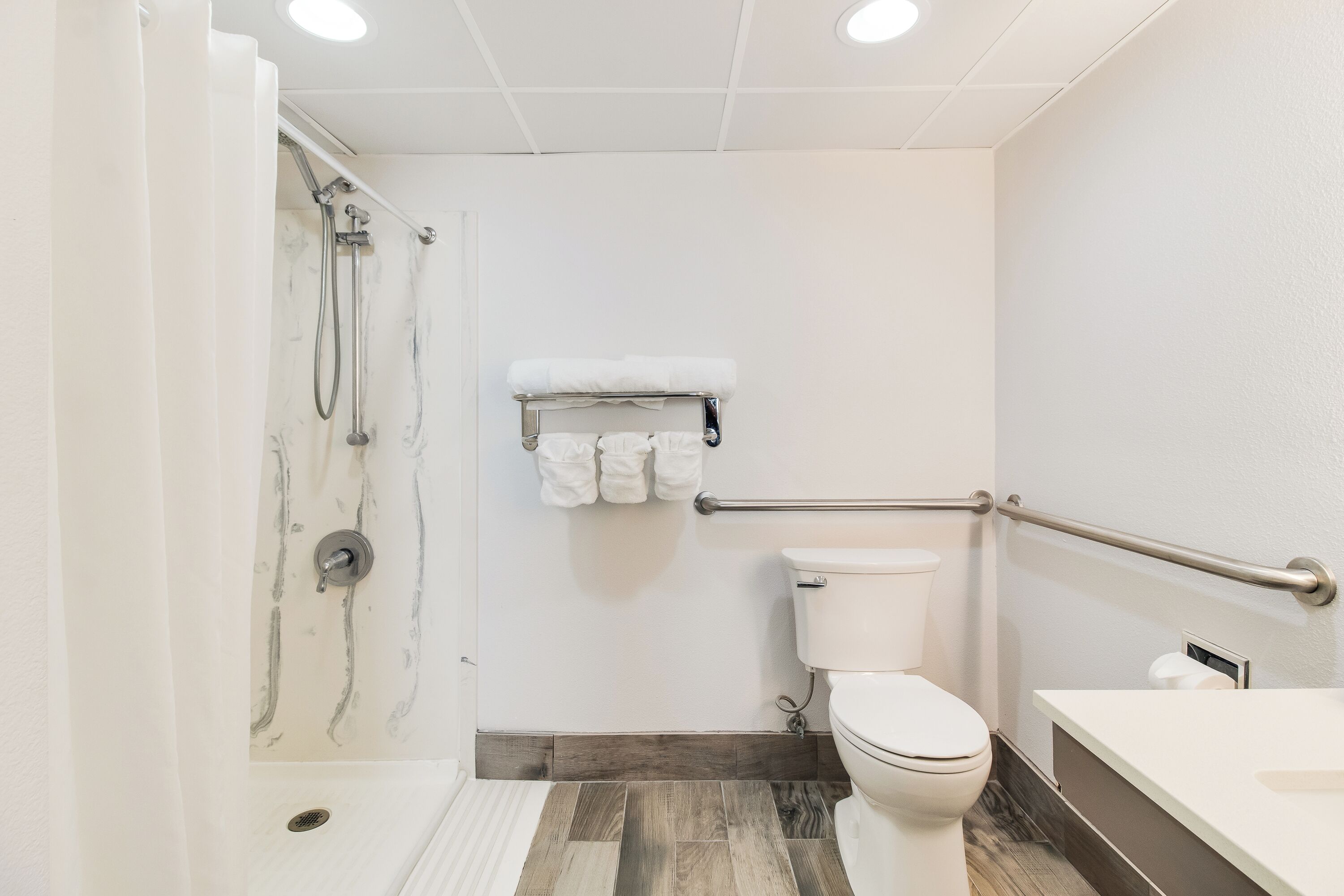 Casa de banho com chuveiro, sanita e lavatório, equipada com barras de apoio e um porta-toalhas.