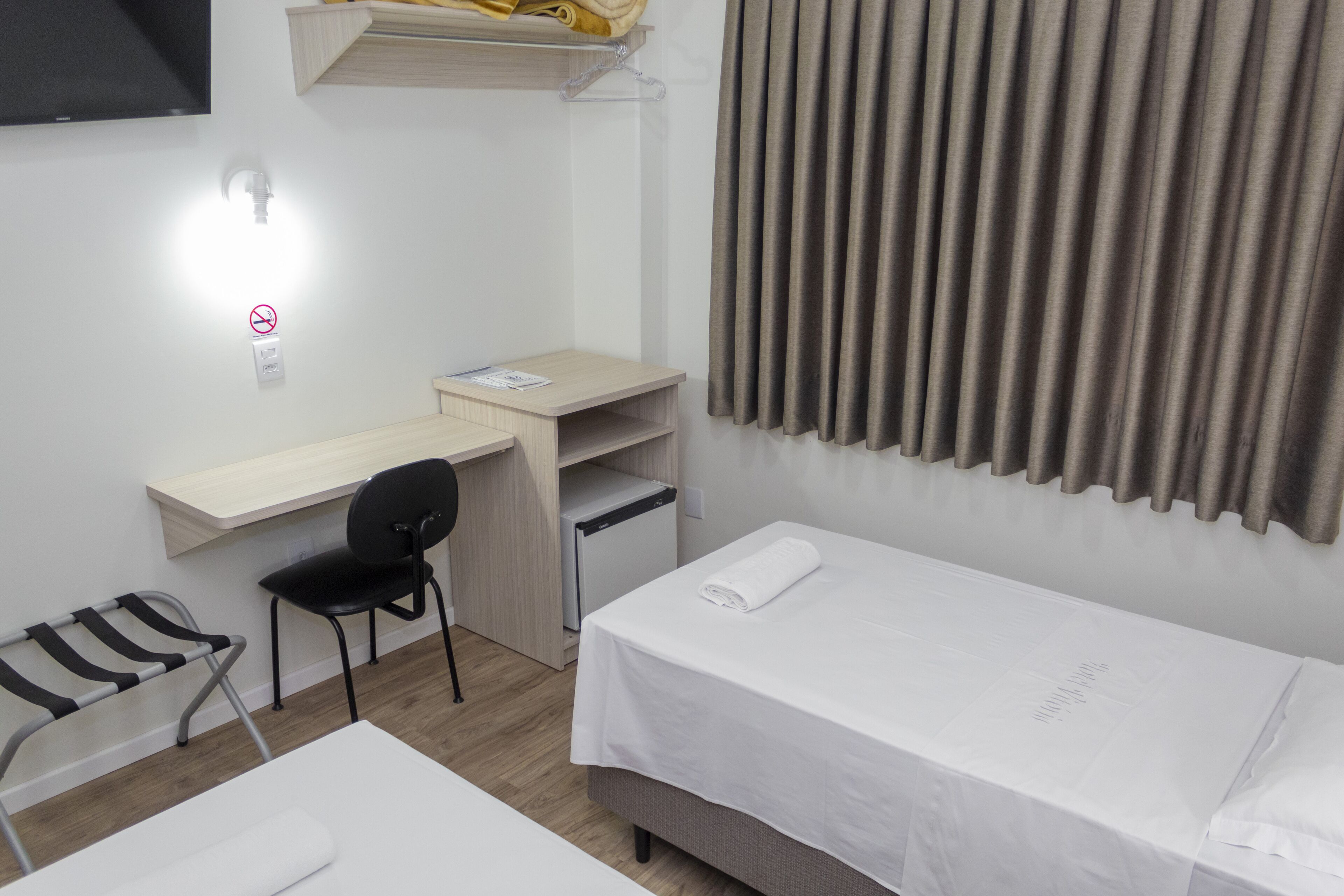 Quarto de hotel com uma cama de solteiro, uma escrivaninha, uma cadeira e uma televisão fixada na parede.