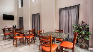 Uma sala de jantar com uma mesa retangular, várias cadeiras de madeira com almofadas laranja, uma televisão de tela plana na parede e um vaso de planta.