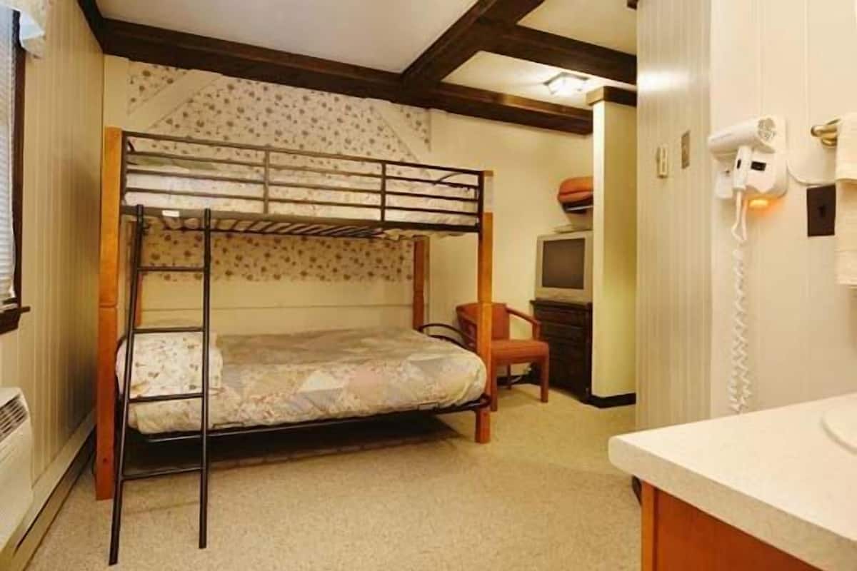 Quarto pequeno e compacto com uma cama beliche, uma secretária pequena com cadeira, um radiador e um secador de cabelo na parede.