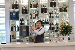 Uma área de bar com um bartender segurando uma garrafa, cercada por prateleiras com diversas garrafas de bebidas e copos.