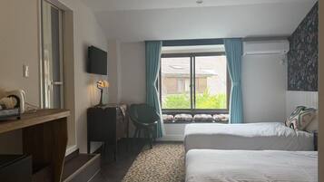 Quarto de hotel com duas camas, uma escrivaninha com candeeiro, uma cadeira e uma janela grande com cortinas.