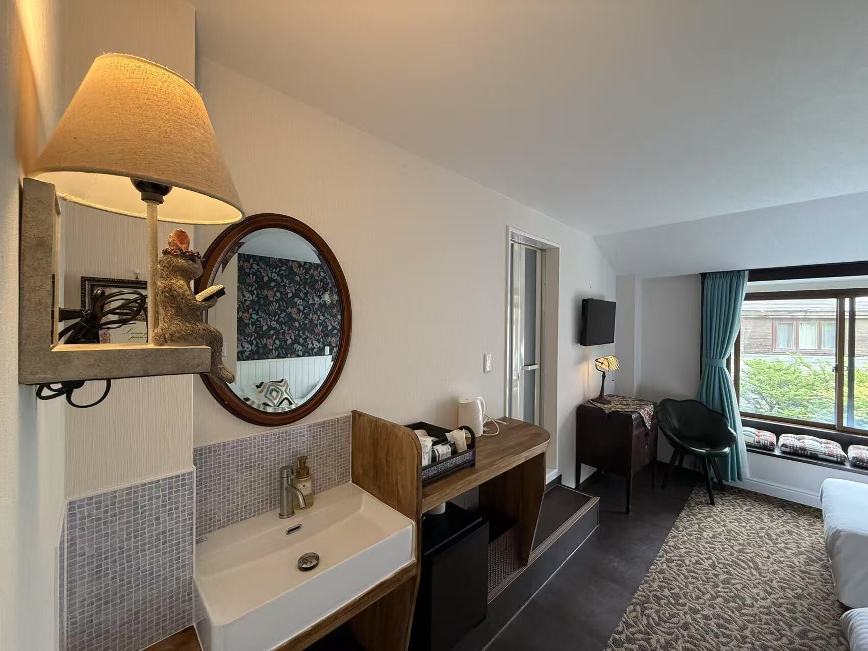 Quarto de hotel com um espelho grande, pia, mesa pequena com abajur, cadeira e televisão fixada na parede.