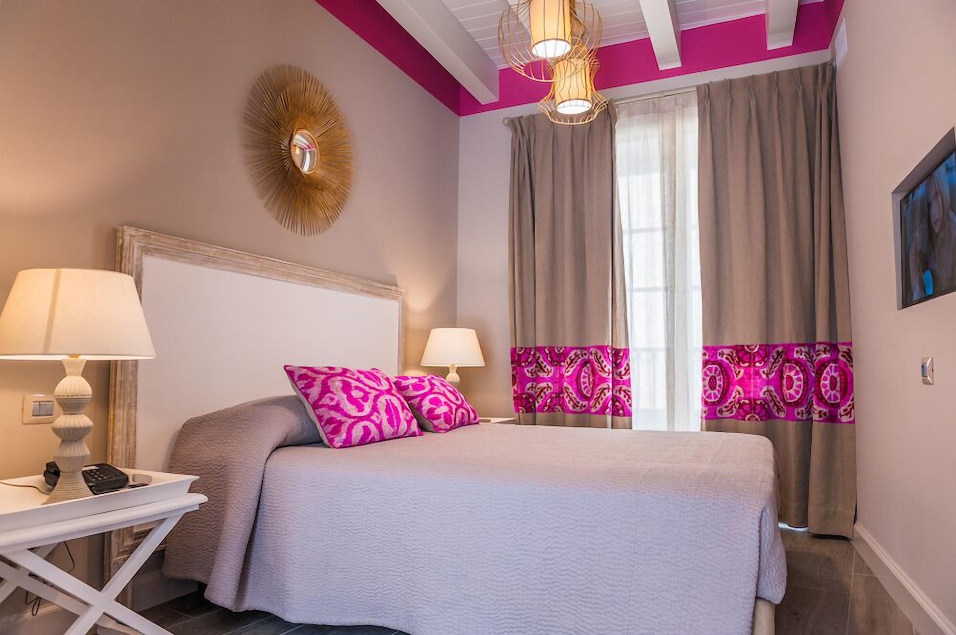 Quarto de hotel com uma cama, duas luminárias laterais, uma mesinha com abajur, um relógio de parede e um espelho grande.